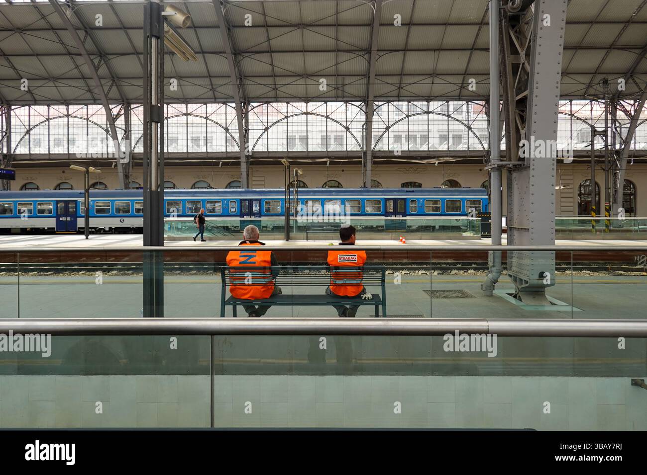 Two railway workers in orange vests sit on a bench facing the tracks ...