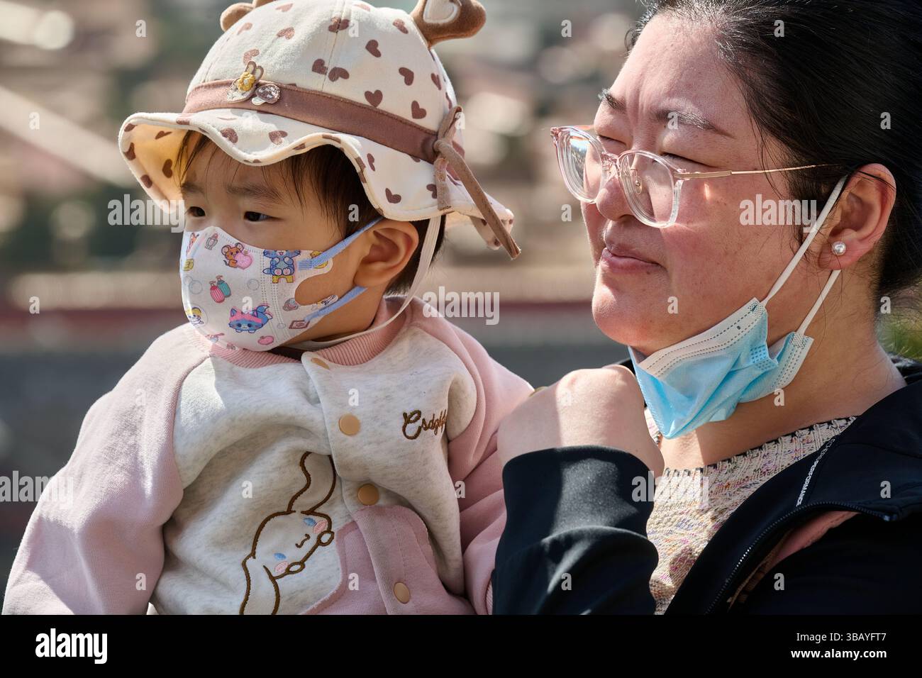 Mother holding a small masked child in patterned clothes and hat, both ...