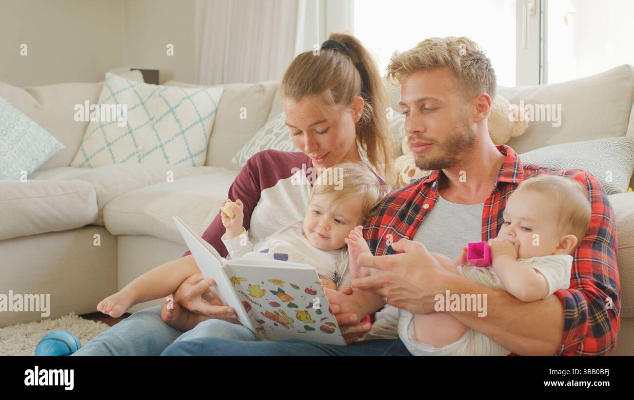 Parents Sit With Young Children On Floor At Home Reading Book Together ...