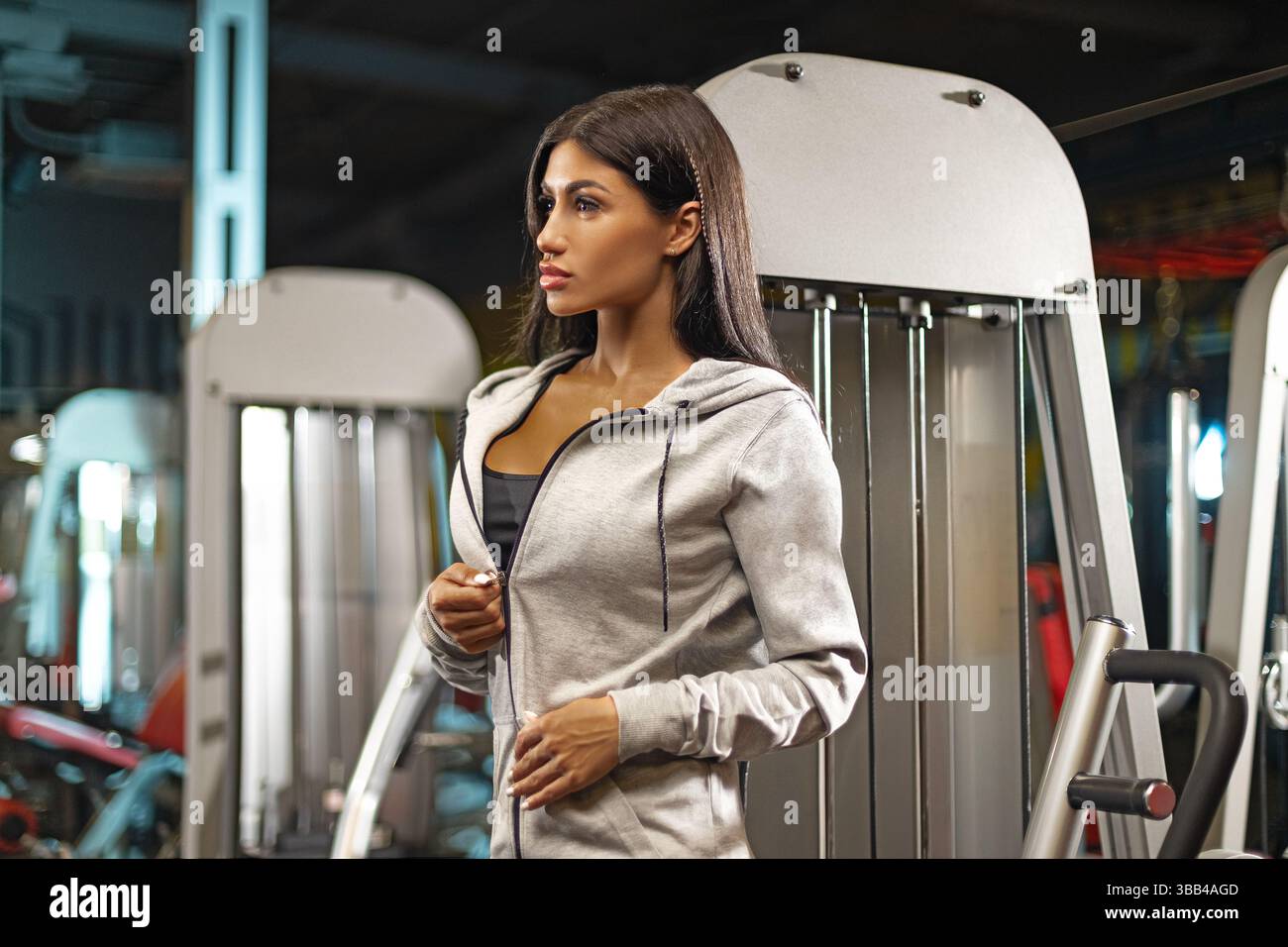 young woman stands in a gym wearing a grey hoodie contemplating her next workout step. The gym ...