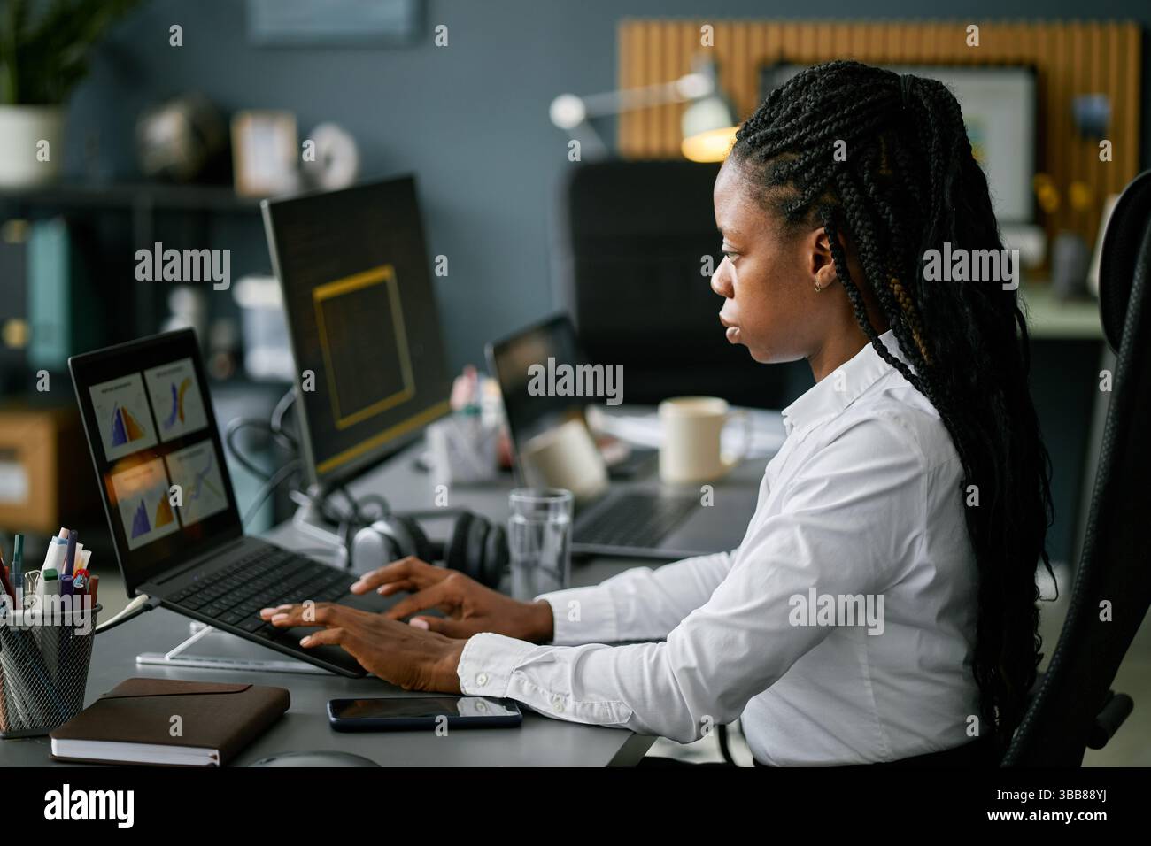 African American woman diligently working at her desk, analyzing data on computer screen while ...