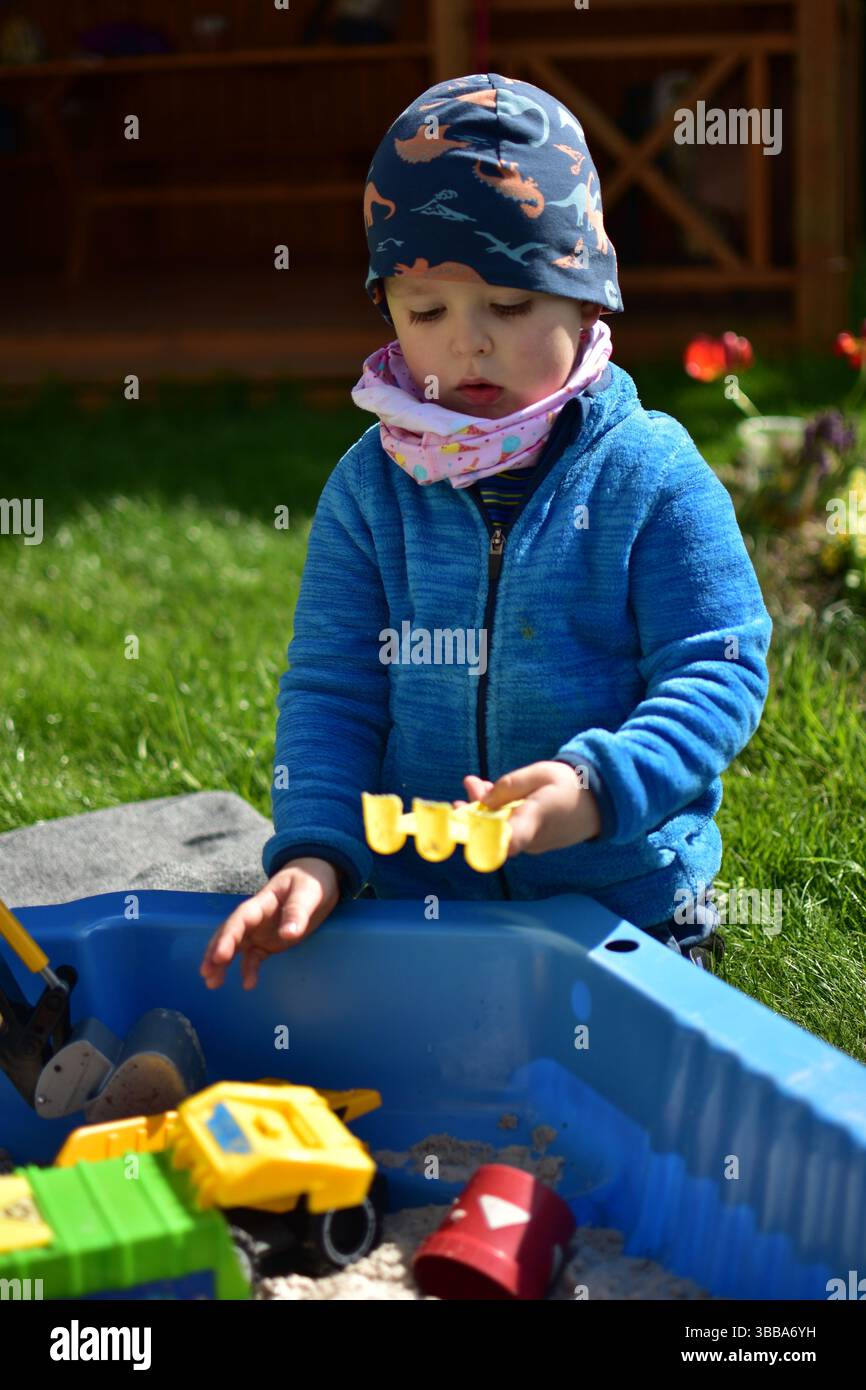 Toddler boy plays with sand toys in a small sandbox in a back yard. Kid ...