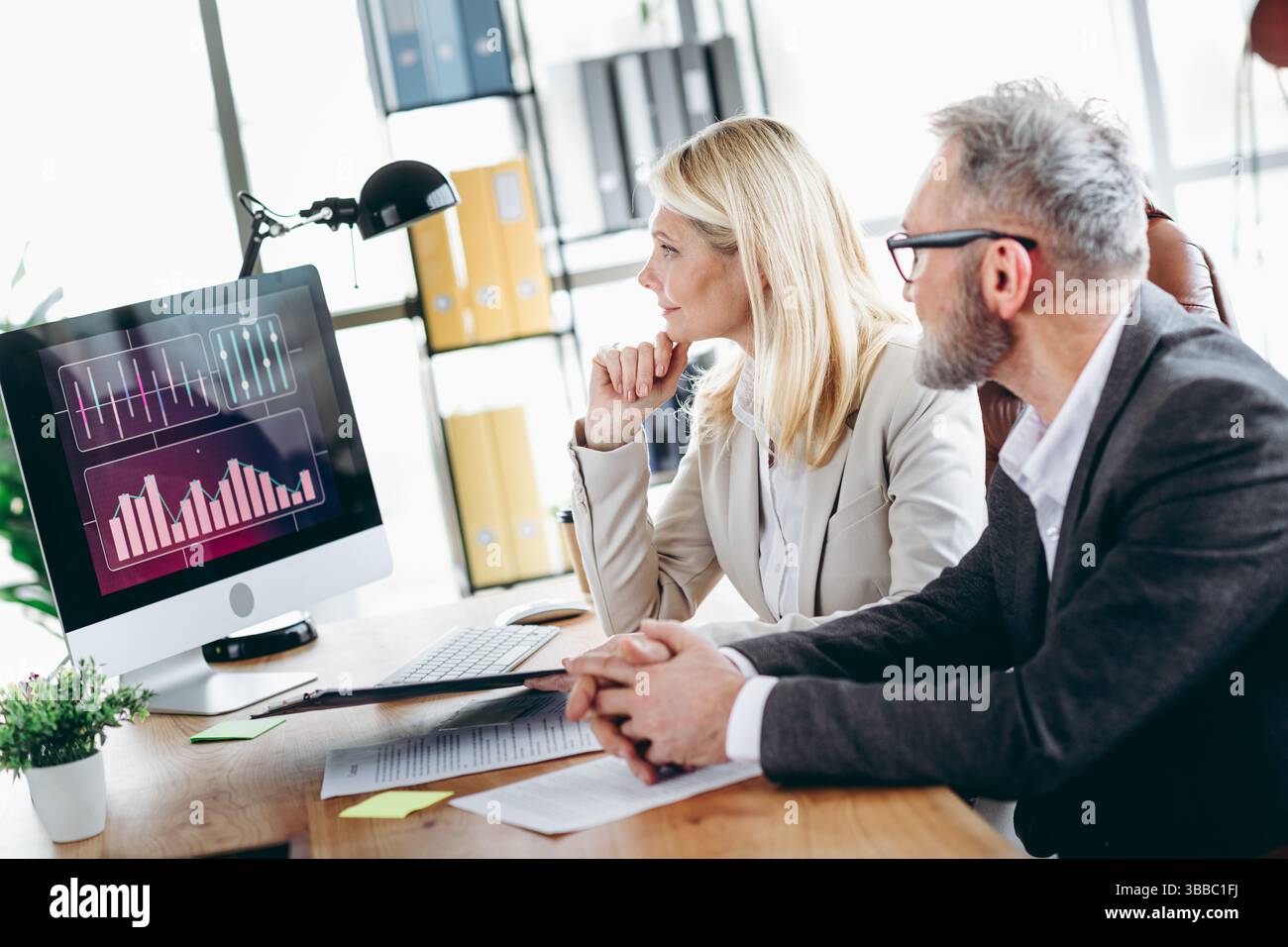 Two professionals analyzing data charts on a computer screen in a modern office setting ...