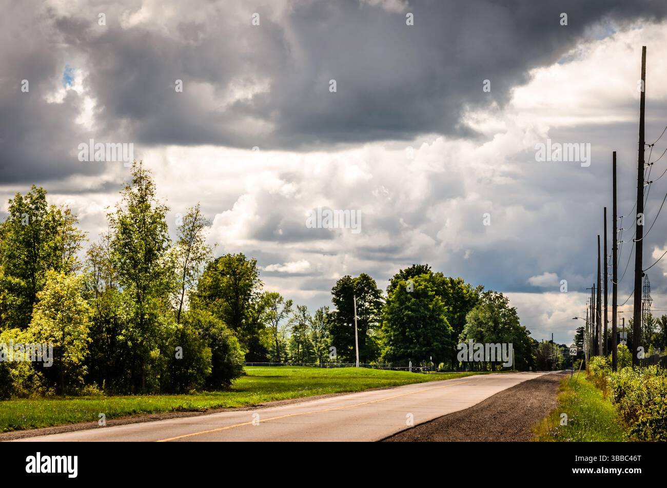 Paved rural road lined with power lines in sunlight with dark clouds ...
