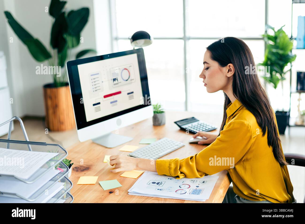 Professional woman working in a modern office setting, analyzing data charts on a desktop ...