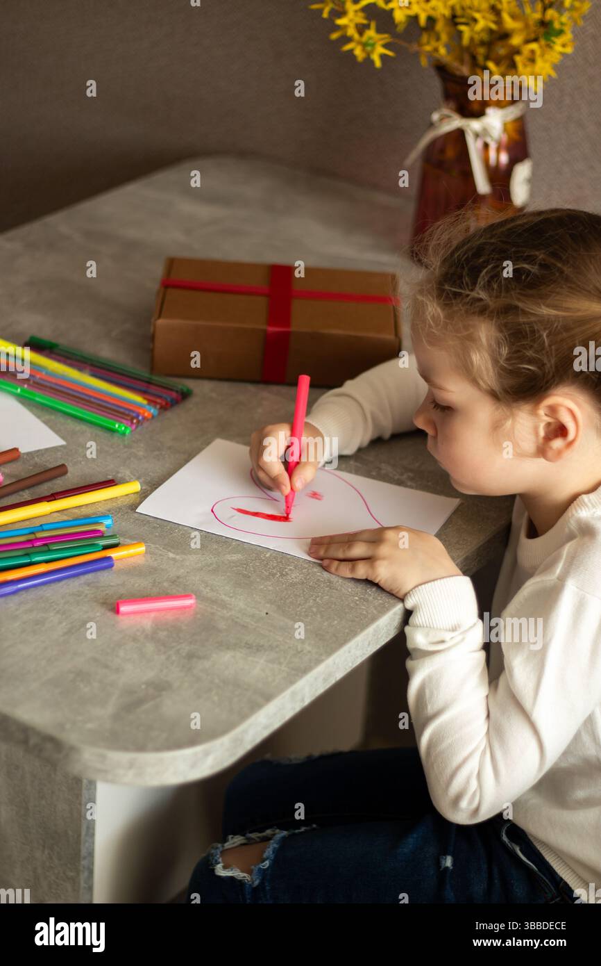 Cute 4-year-old blonde girl concentrates while drawing sitting at table ...