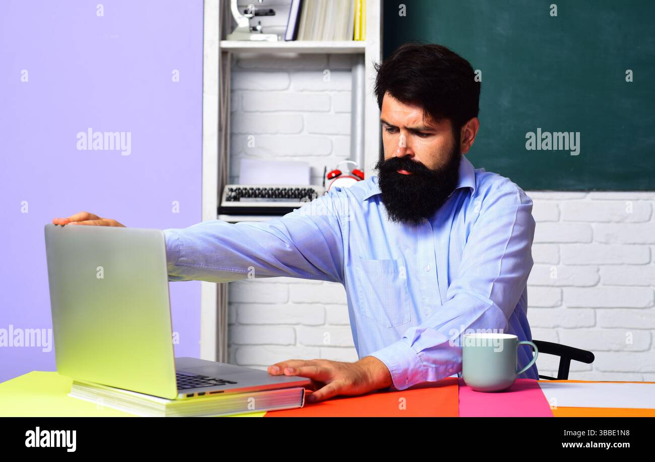 Back to school. Serious male teacher sitting at desk in classroom, preparing for university exam ...