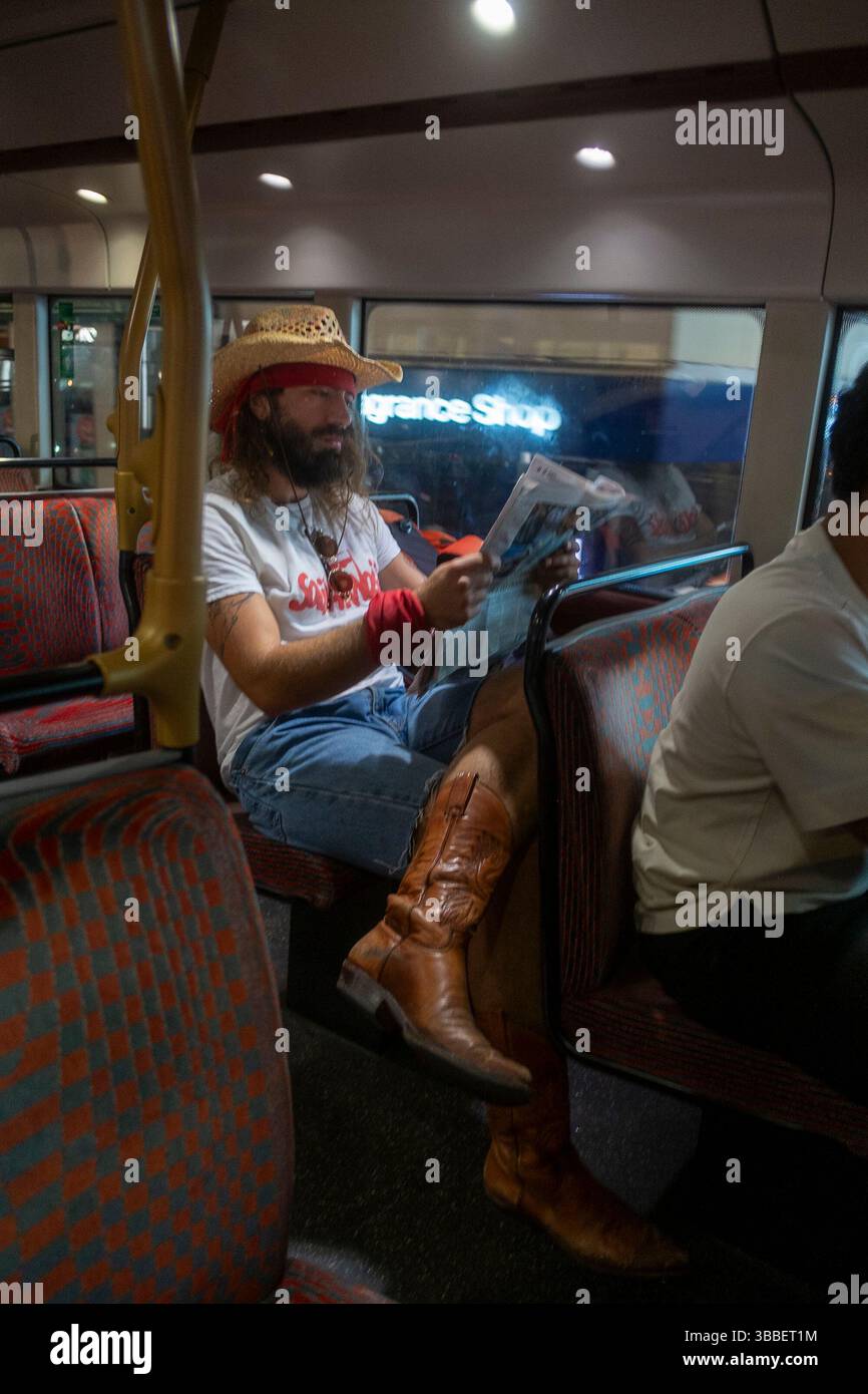 Man wearing cowboy boots and straw hat reads newspaper on London night ...