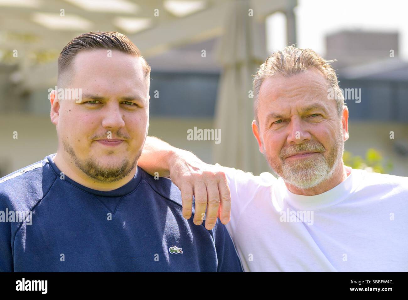 A father and adult son stand closely side by side in natural light ...
