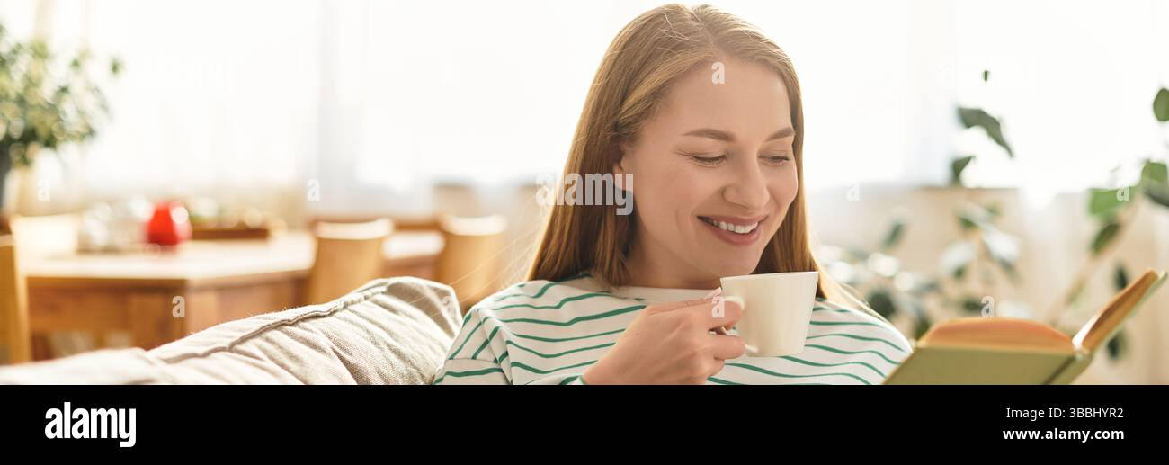 A young woman smiles as she sips her warm drink while reading a book in ...