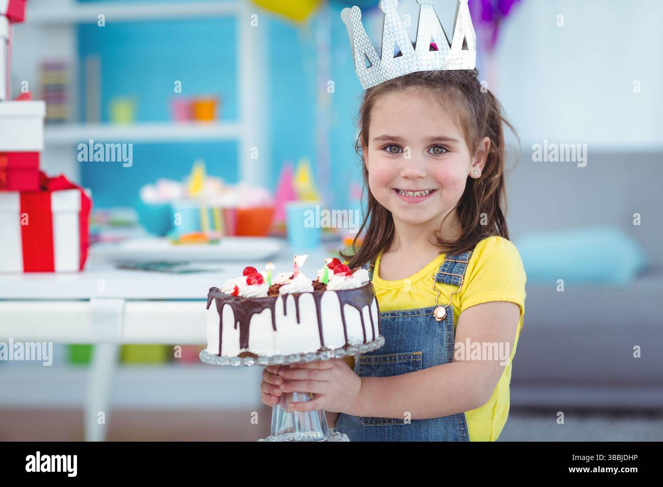 Smiling child wearing paper crown holding frosted cake and gift boxes at birthday party, copy ...