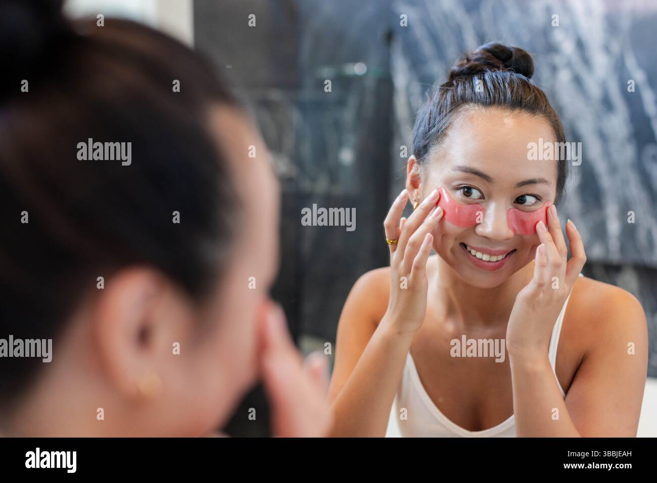 Asian woman pressing pink under-eye gel patches at vanity mirror, with ...