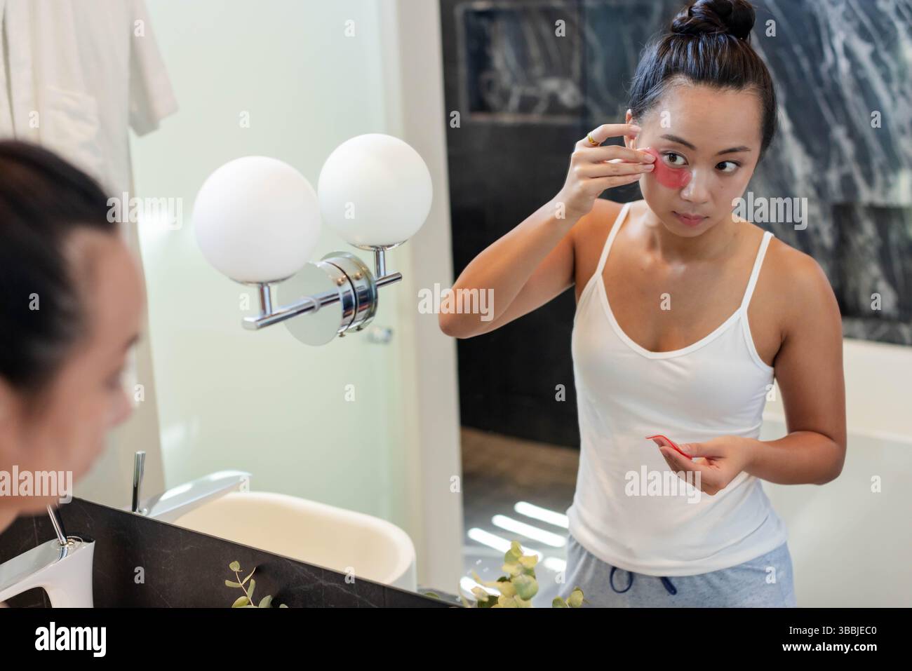 Standing Asian woman applying red hydrogel under-eye patch at bathroom ...