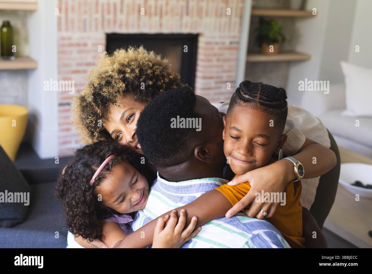 Embracing African American family sitting on gray sofa in living room, with brick fireplace ...