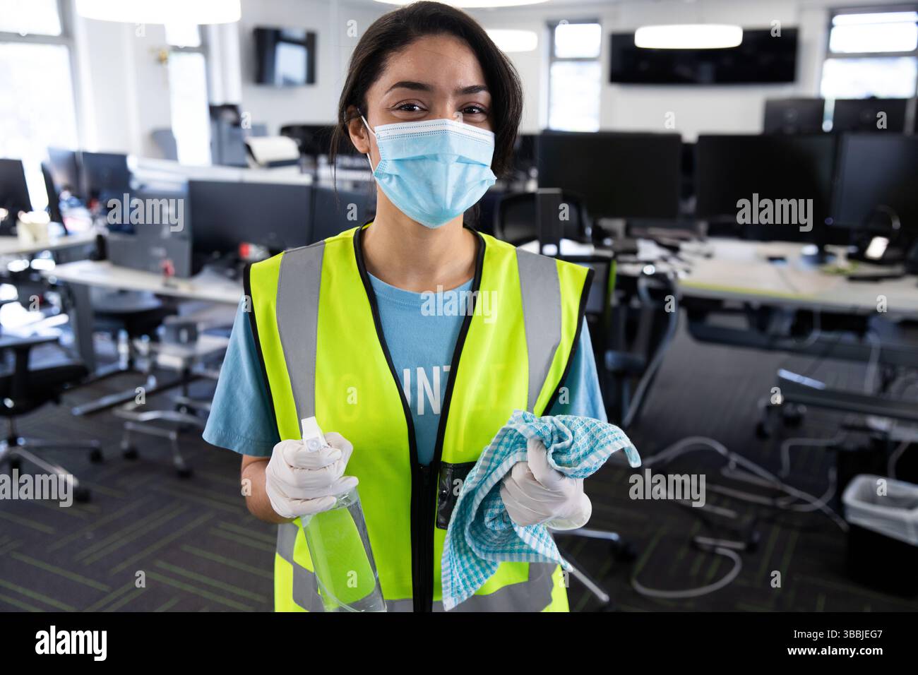 Wiping desks in modern office, woman wearing safety vest and gloves ...