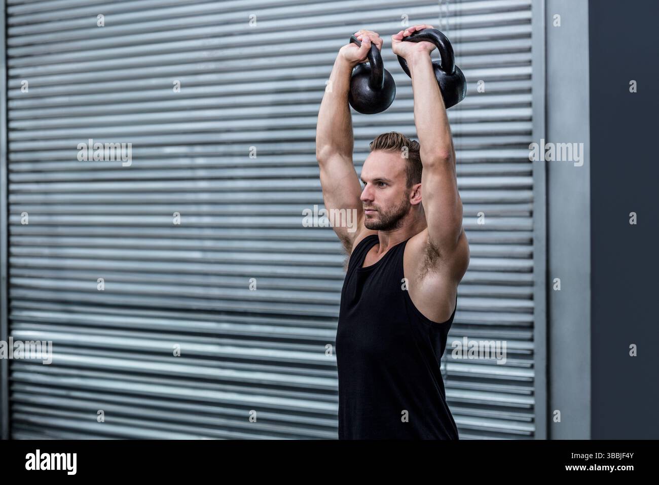 Lifting athletic man pressing two kettlebells overhead before garage ...