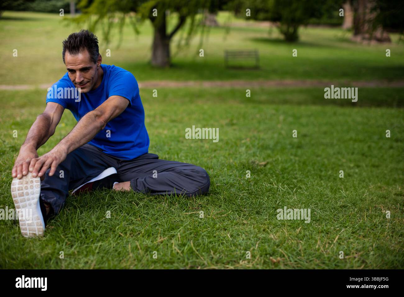 Stretching man grasping extended leg on grass in public park, with ...