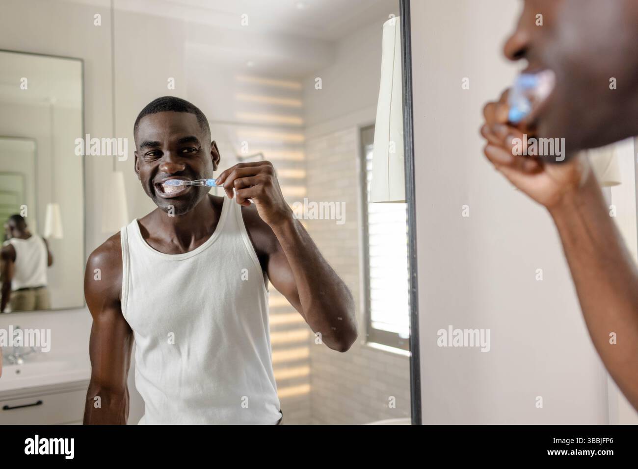 African American man brushing teeth in bathroom at sink, with mirror reflection and toothbrush ...