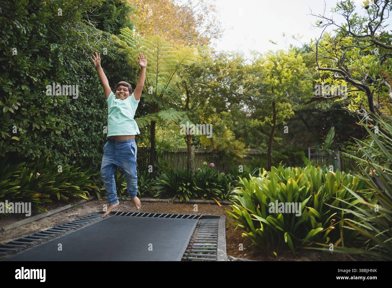 Boy jumping on in-ground trampoline in lush backyard garden, surrounded ...