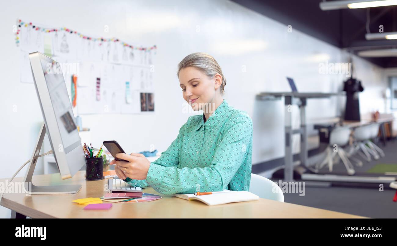 woman scrolling smartphone at desk in open office, with computer ...