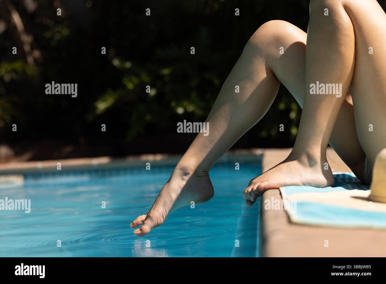 Sitting Asian woman dipping foot in pool water on tiled deck, with swimsuit bottom, copy space ...