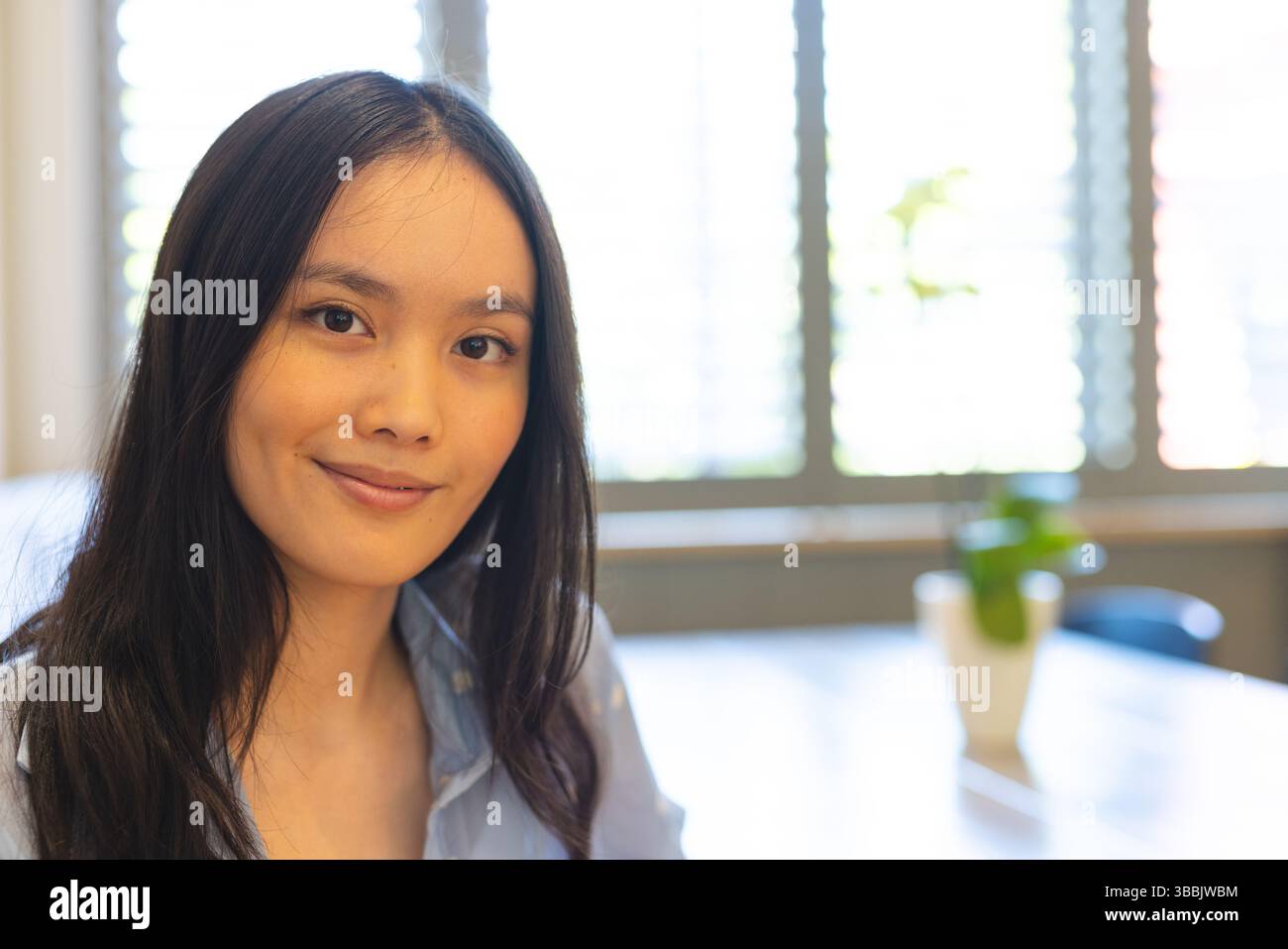Sitting Asian woman looking at camera at light wood study table, potted ...