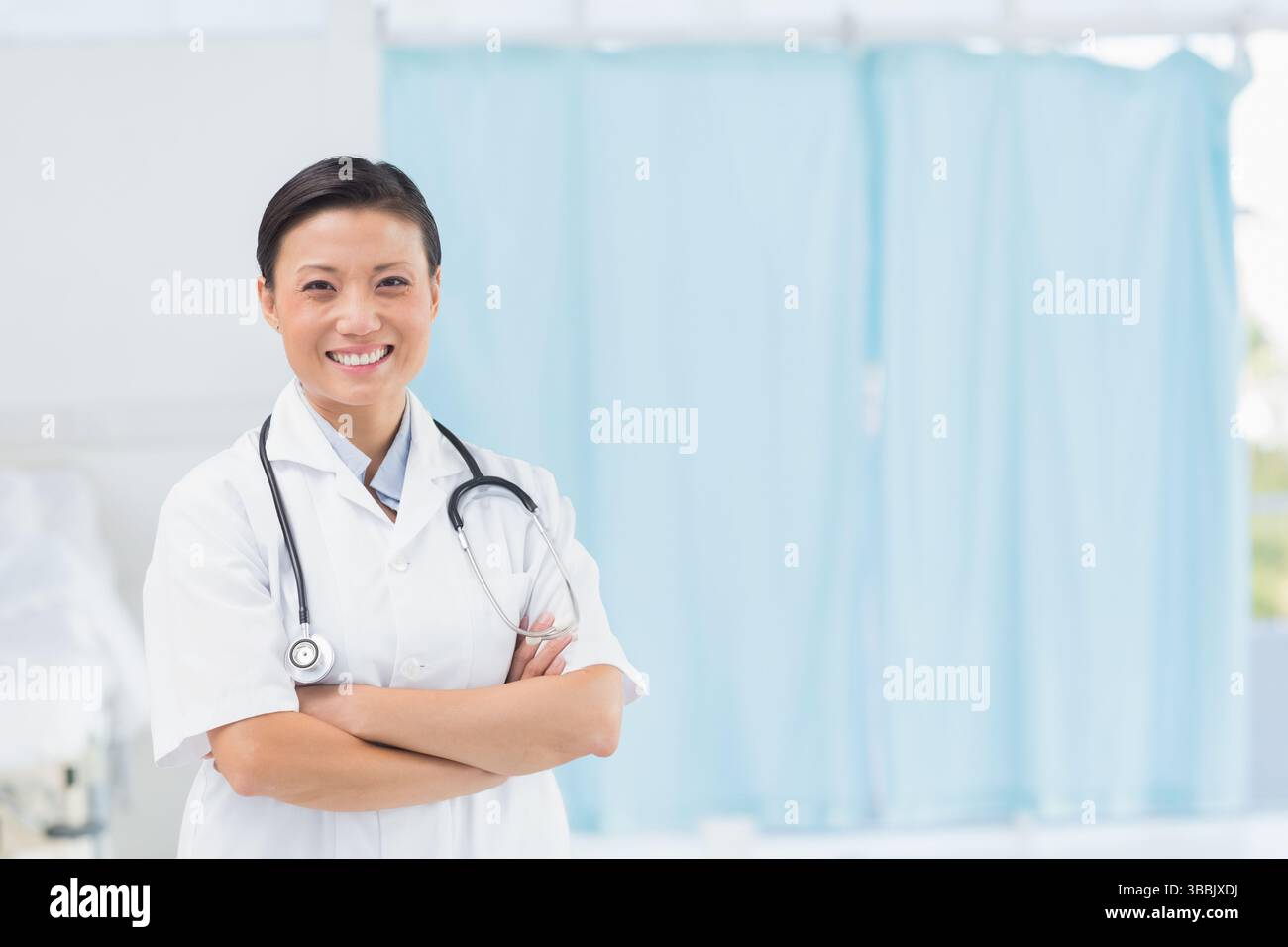 Smiling Chinese female doctor wearing lab coat with stethoscope in exam room with bed, copy ...