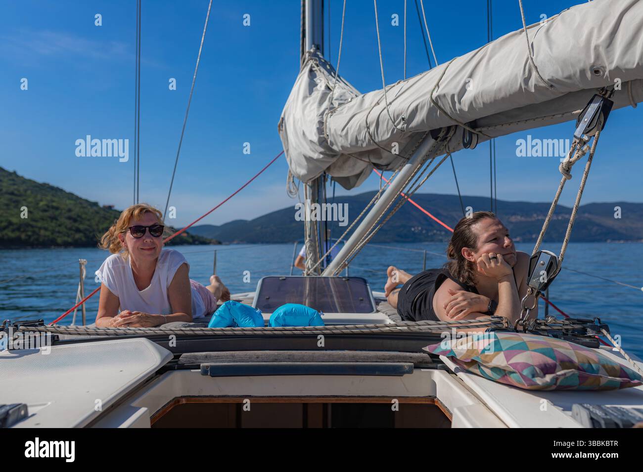 Man and woman lying on padded deck space of a sailing yacht, shaded by mast and surrounded by ...