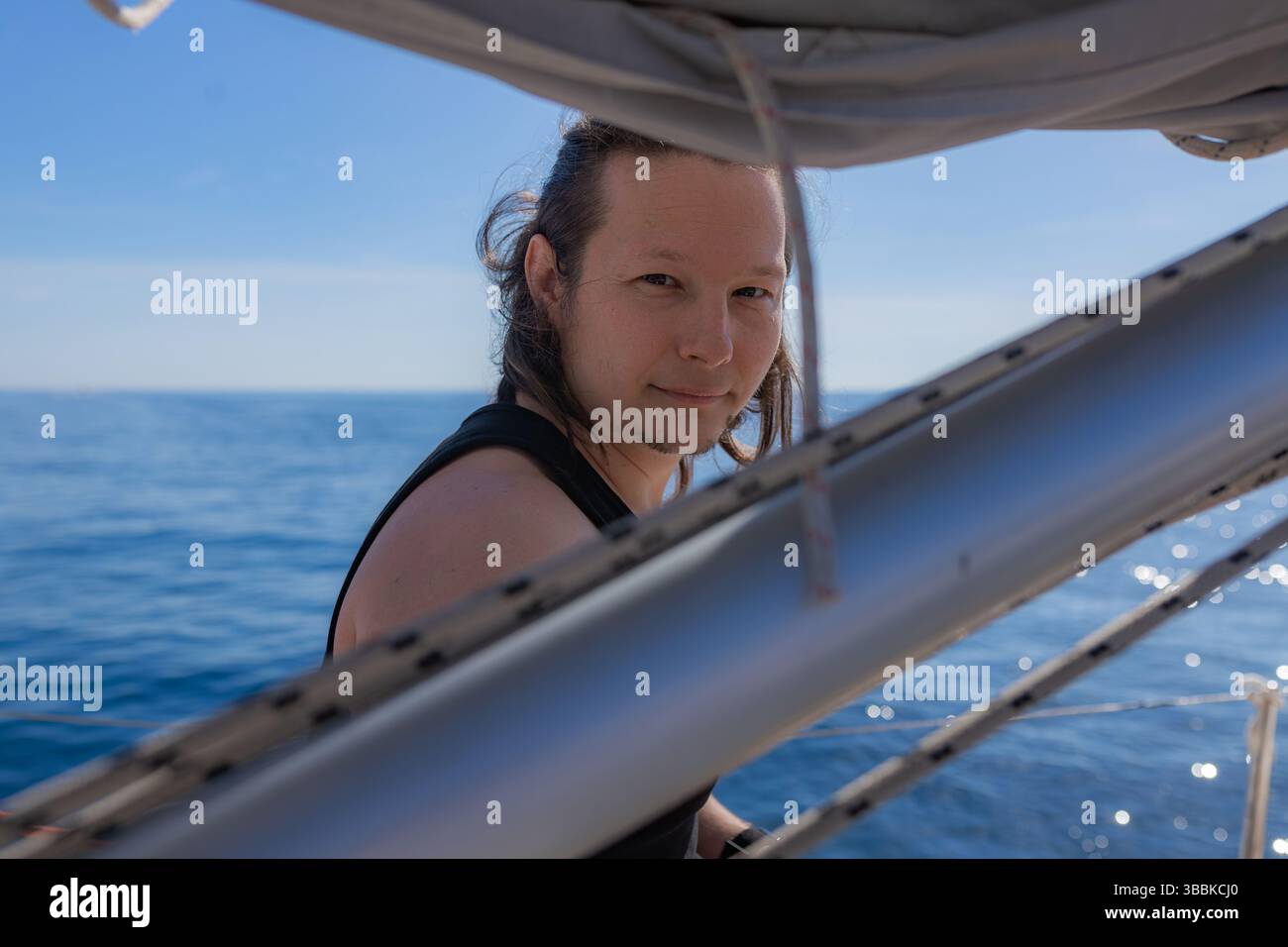 Young man smiling softly, framed by yacht rigging, blue ocean horizon ...