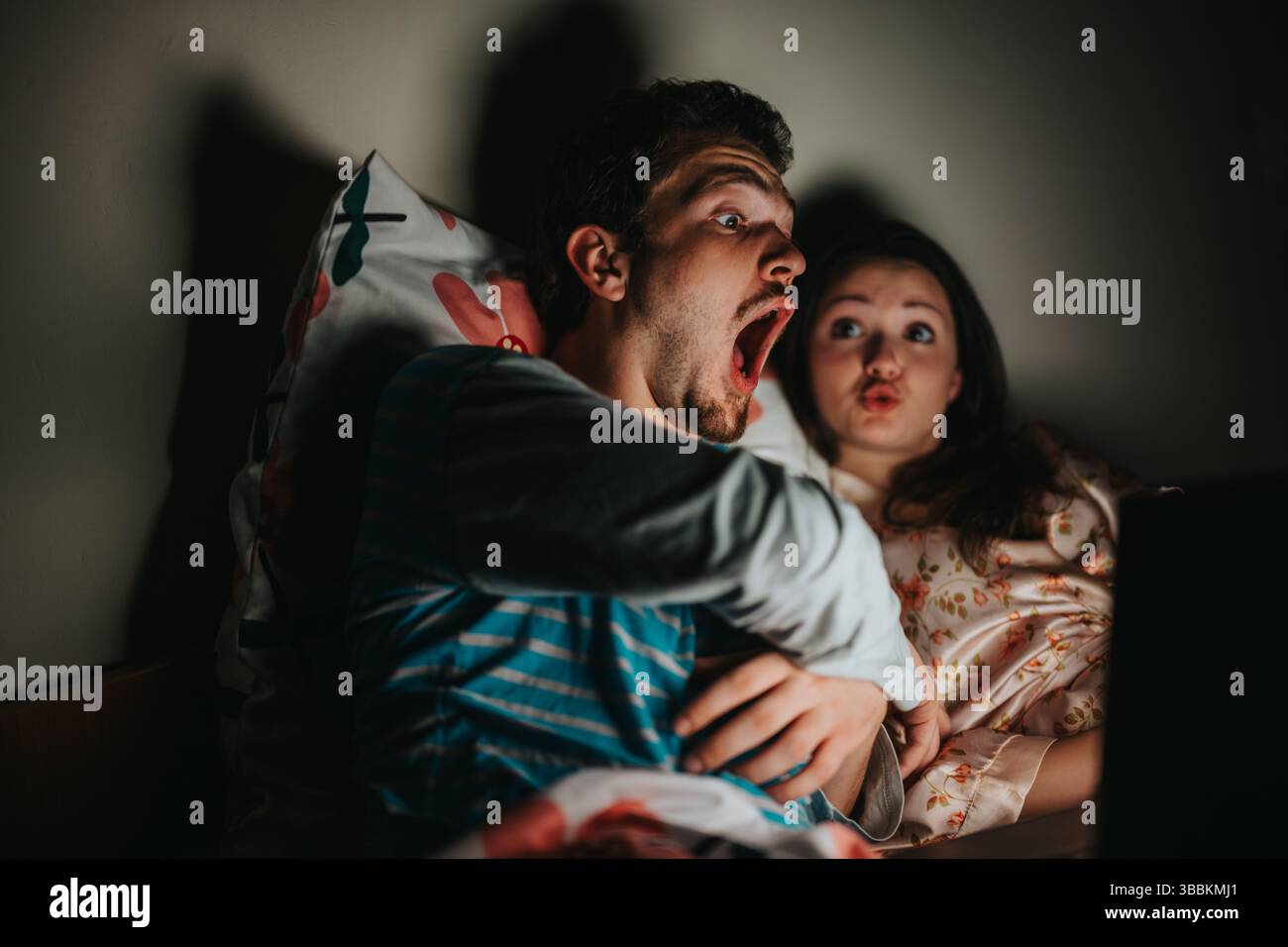 Couple watching a movie with surprised expressions in bed at night ...