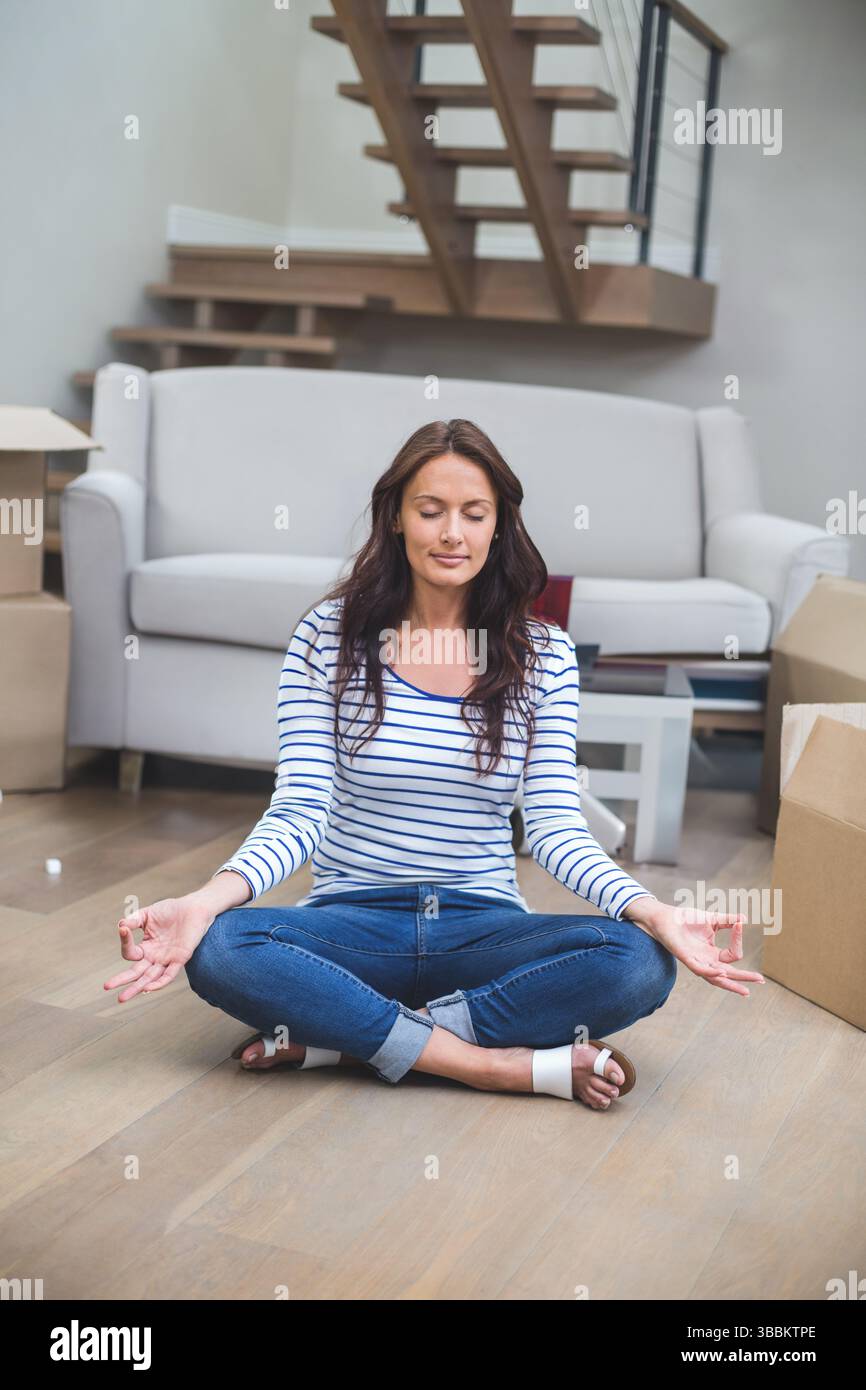 Meditating woman sitting cross-legged on hardwood floor in modern living room, with moving boxes ...