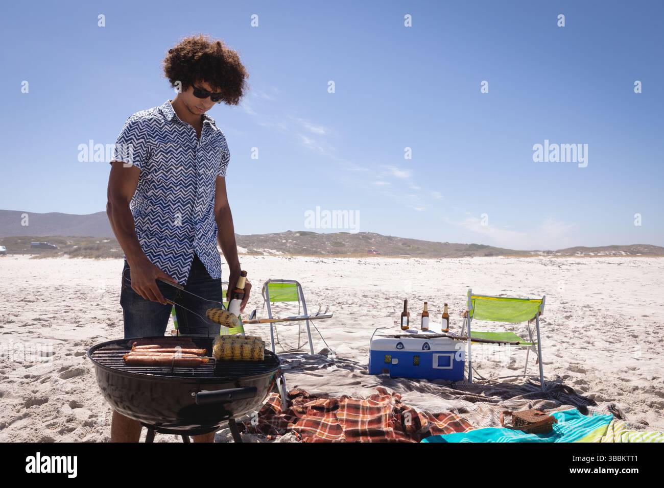 Grilling man turning corn and sausages with metal tongs on sandy beach ...