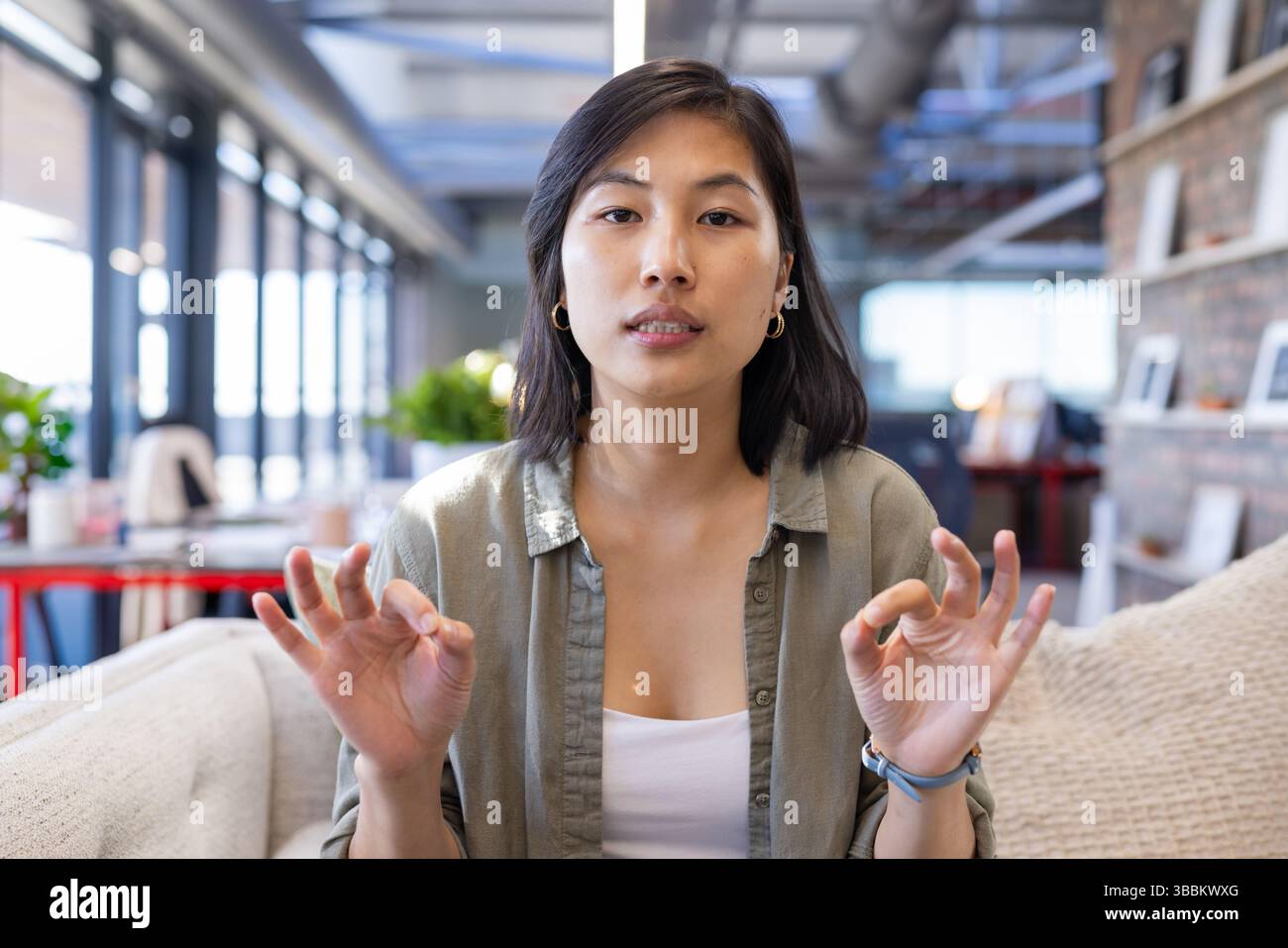 Young Asian woman making OK gesture while sitting on couch in modern office, showing confidence ...