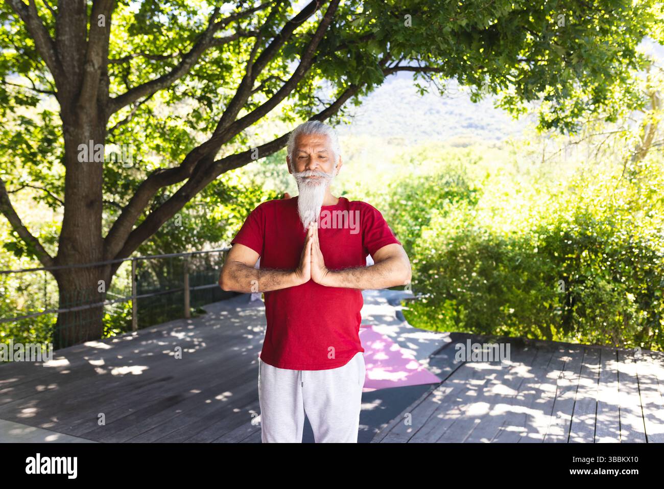 Senior man praying on wooden deck under tree canopy with rope railing ...
