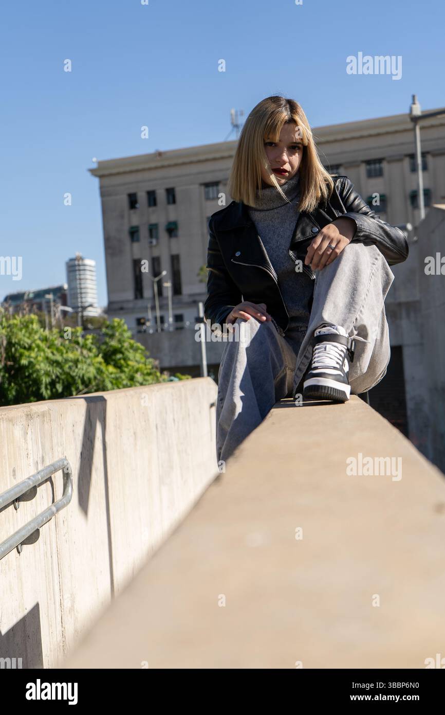 Young stylish woman sitting on a wall in urban setting, enjoying sunny ...
