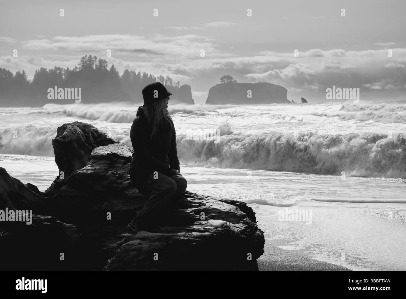 A woman sits on driftwood gazing out at crashing waves and sea stacks ...