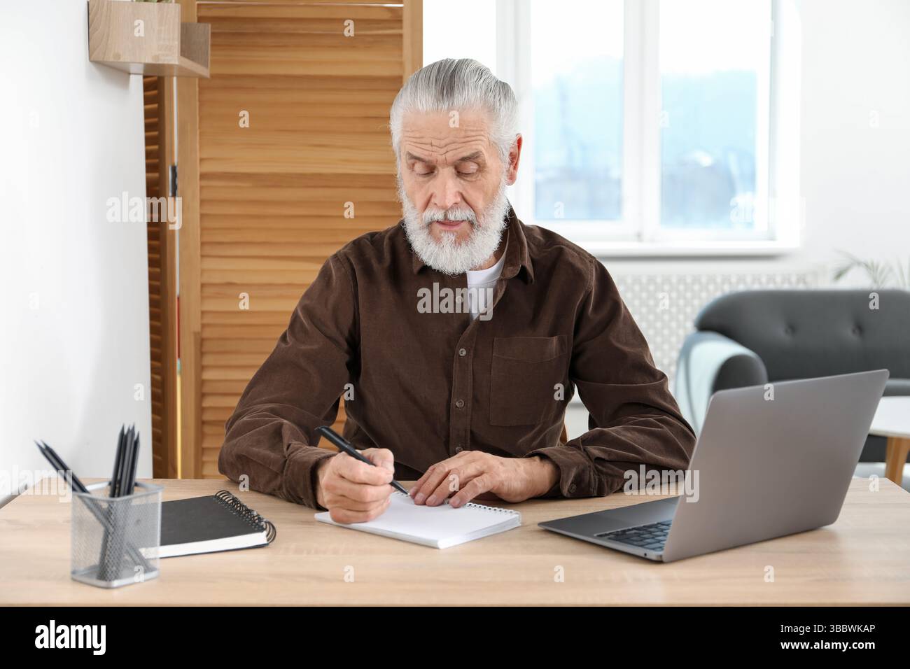 Senior man taking notes while learning online at desk indoors. Self ...