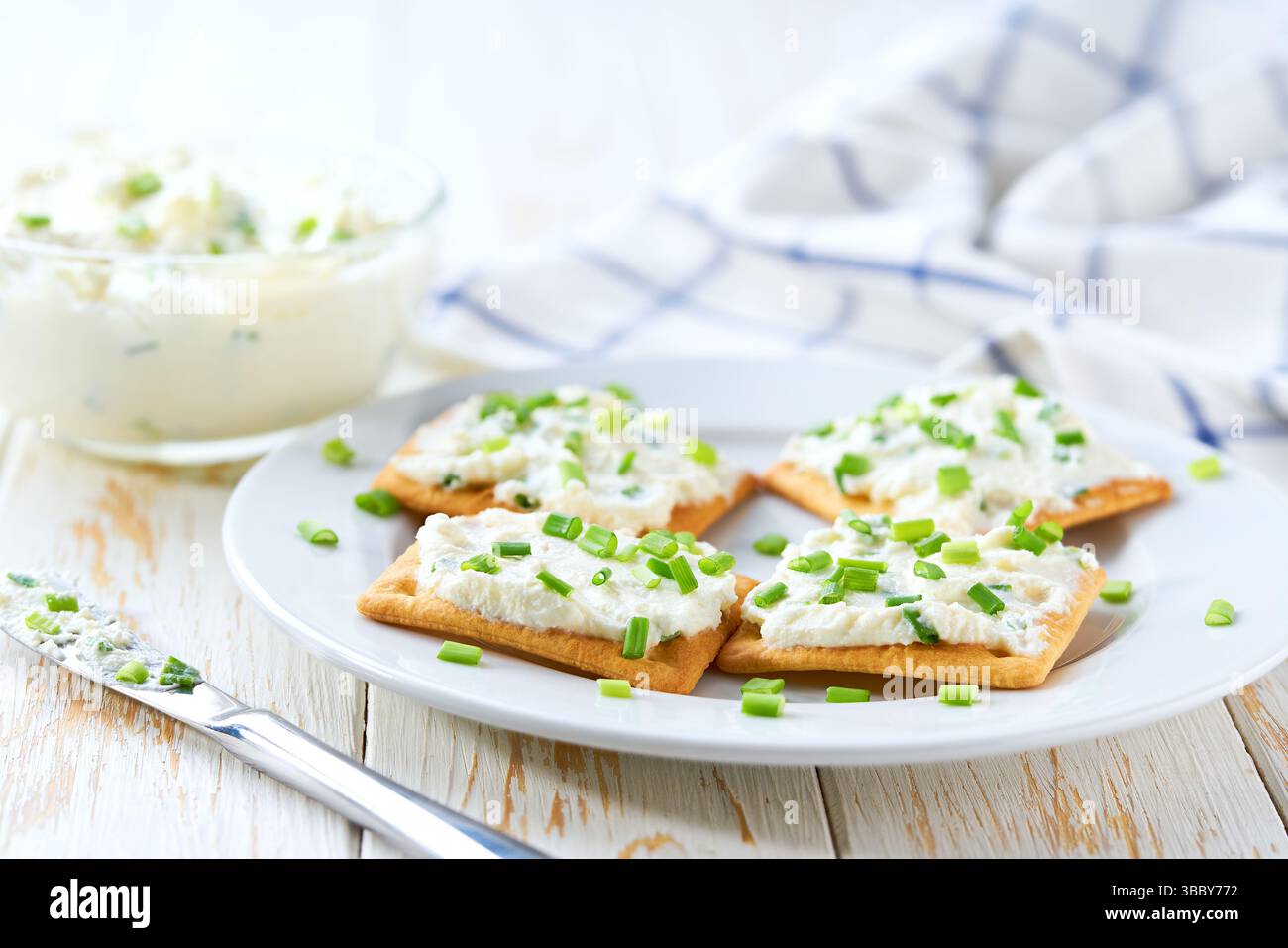 Crispy crackers and a glass bowl with a cream cheese spread and cut chives on a light kitchen ...