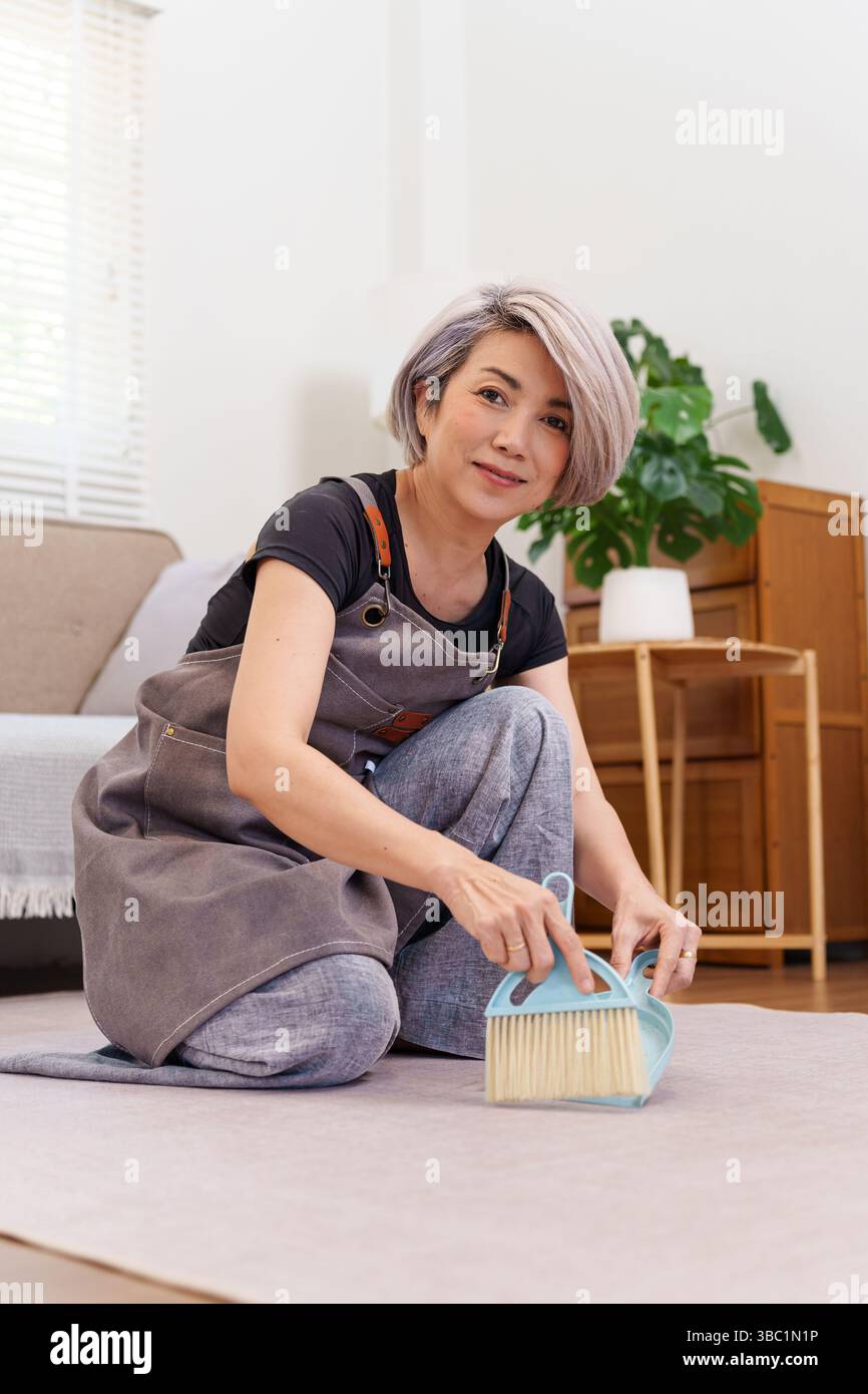 An Asian older woman is happily cleaning her home floor with a broom ...