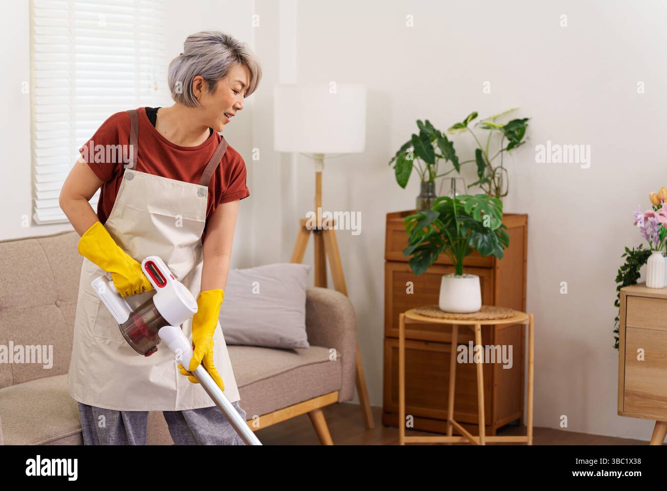 A 40-50 year old Asian woman uses a cordless vacuum cleaner in her modern home. She wears gloves ...