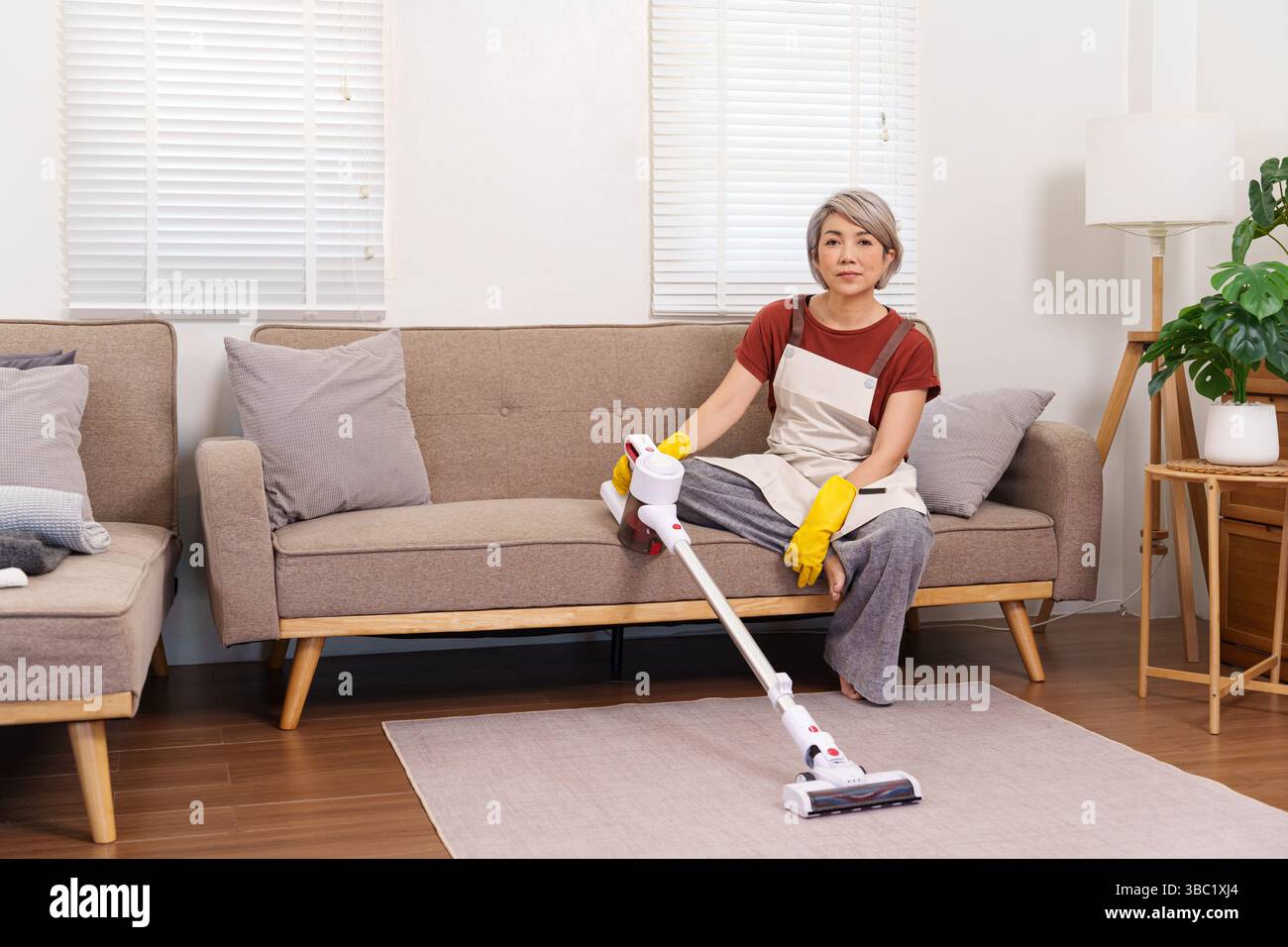Middle-aged Asian woman in apron vacuuming living room with handheld ...