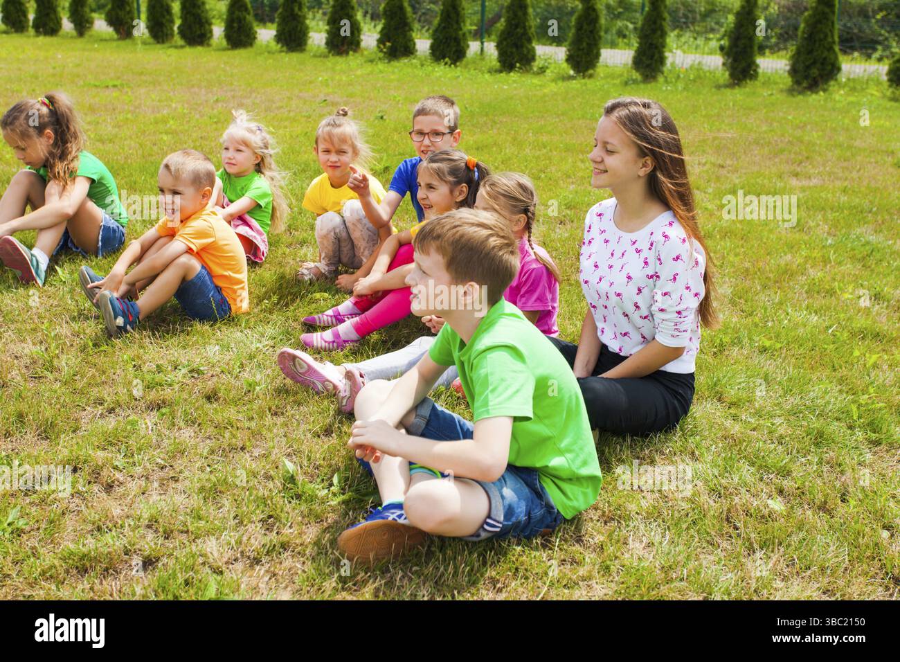 Group of smiling children sitting on a grassy ground, listening with ...