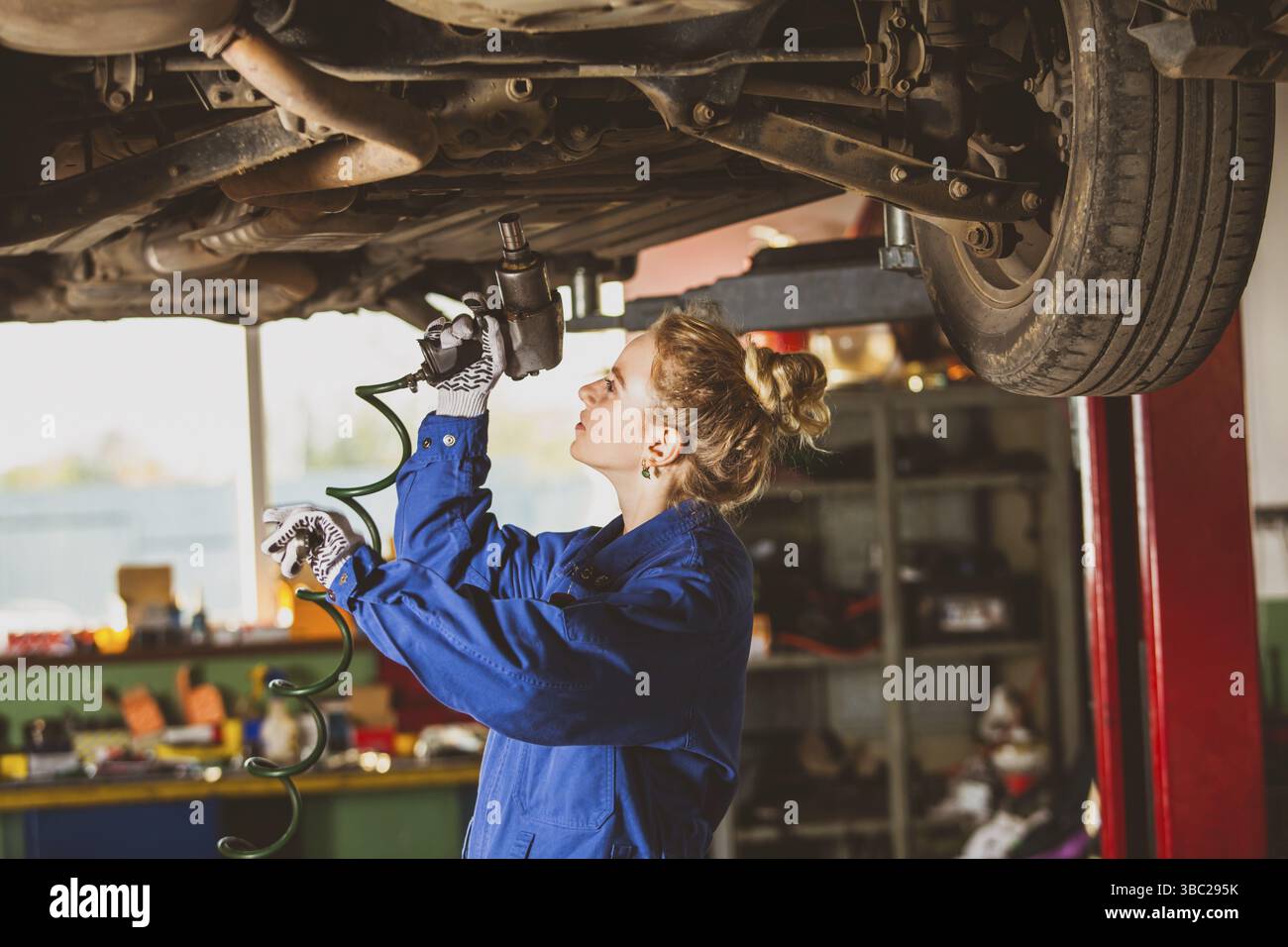 Young female mechanic with blonde hair in blue overall, working under ...