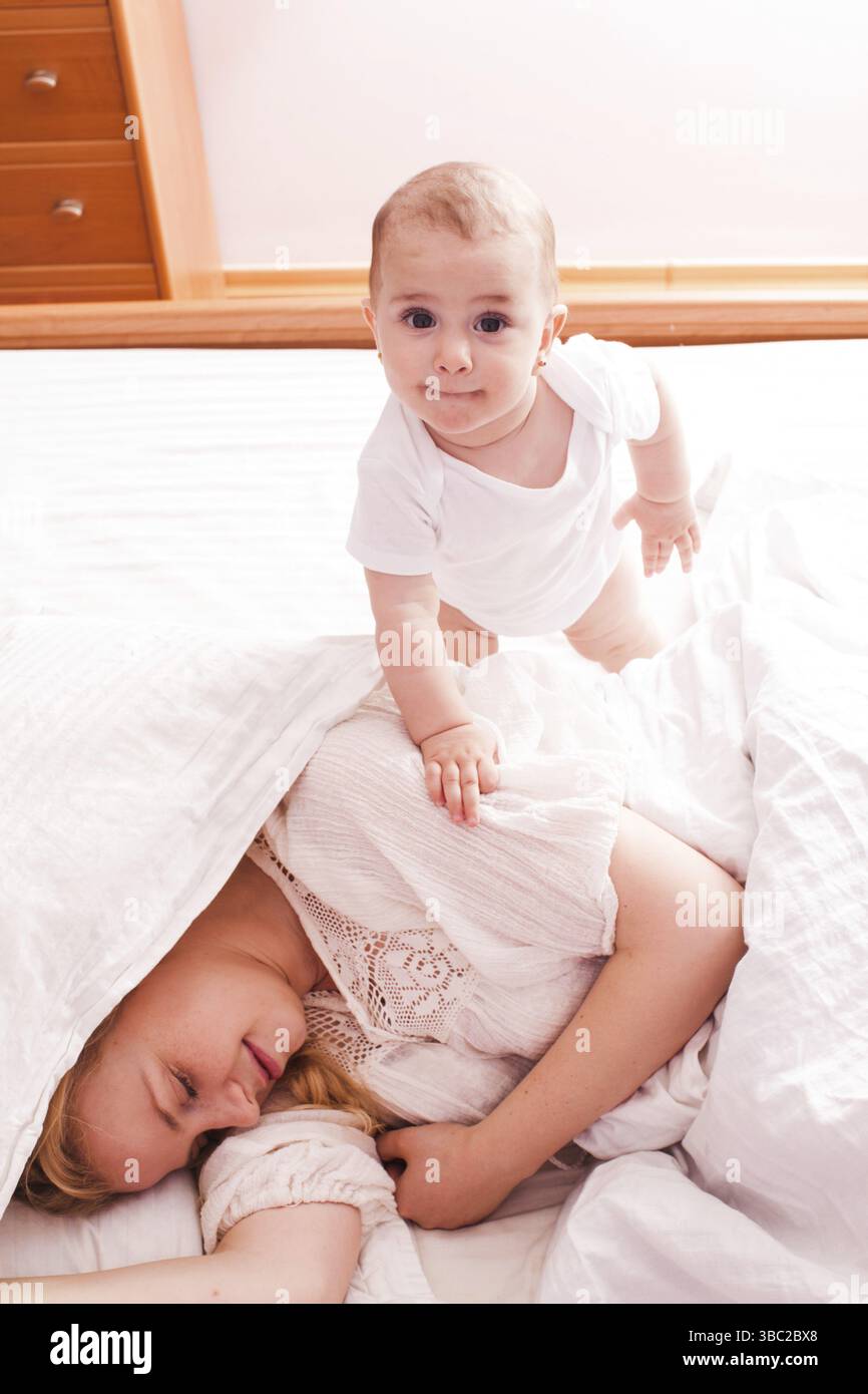 Cute Baby Girl Came To Wake Sleepy Mom Which Sleeping On A White Bed