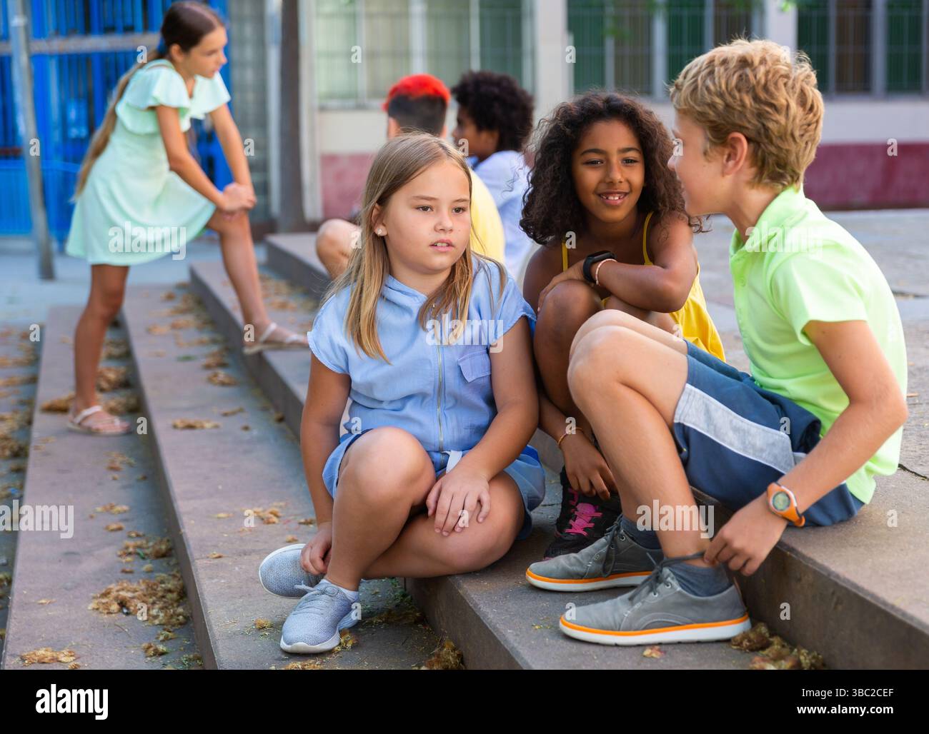 Cheerful preteen girls and boys sitting on steps outdoors and chatting ...