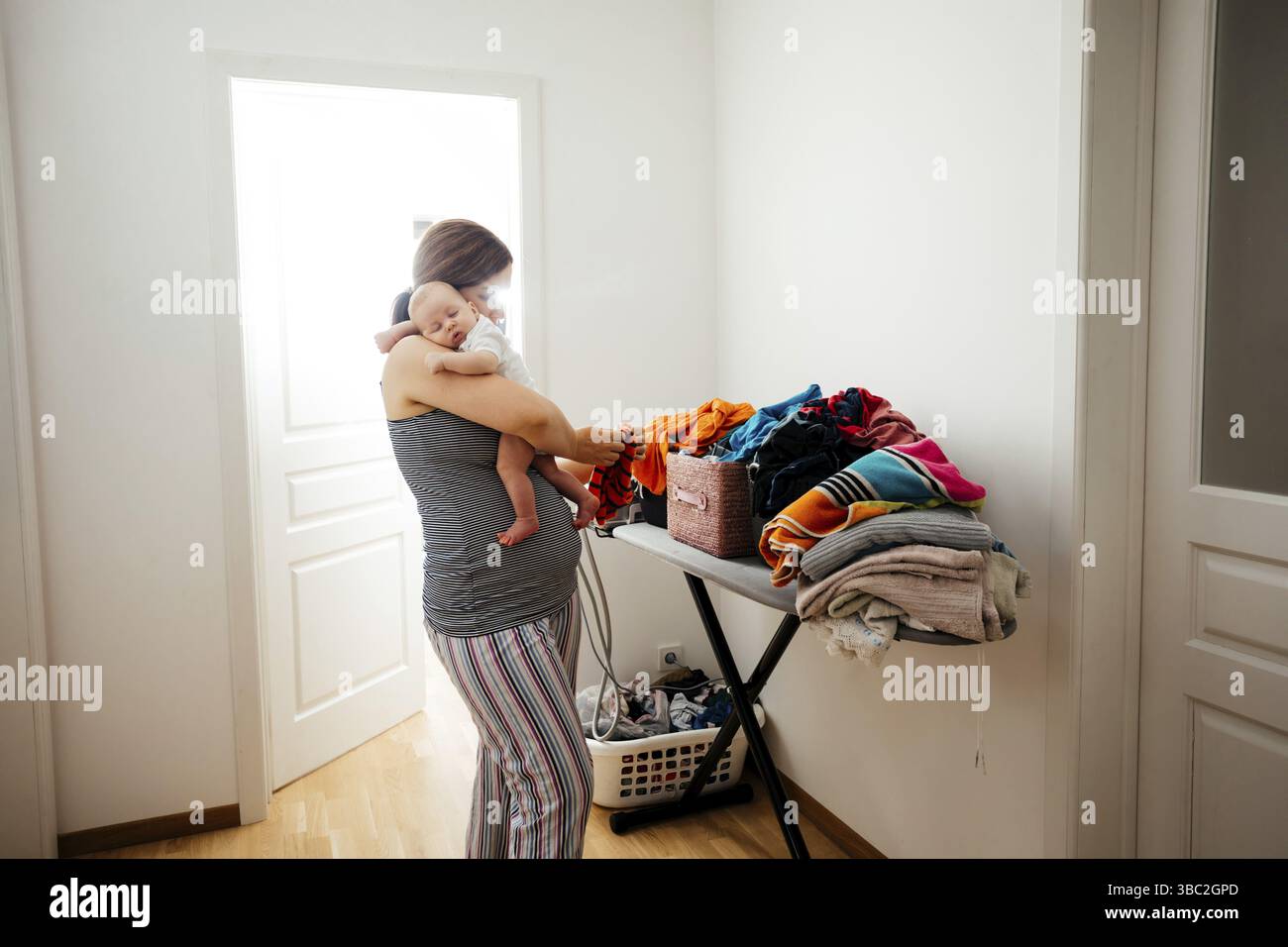 Young mother and newborn baby together engaged in housework preparing laundry for ironing ...