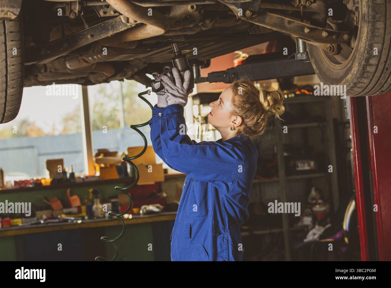 Side view of young blonde woman in blue coverall, fixing car on ...