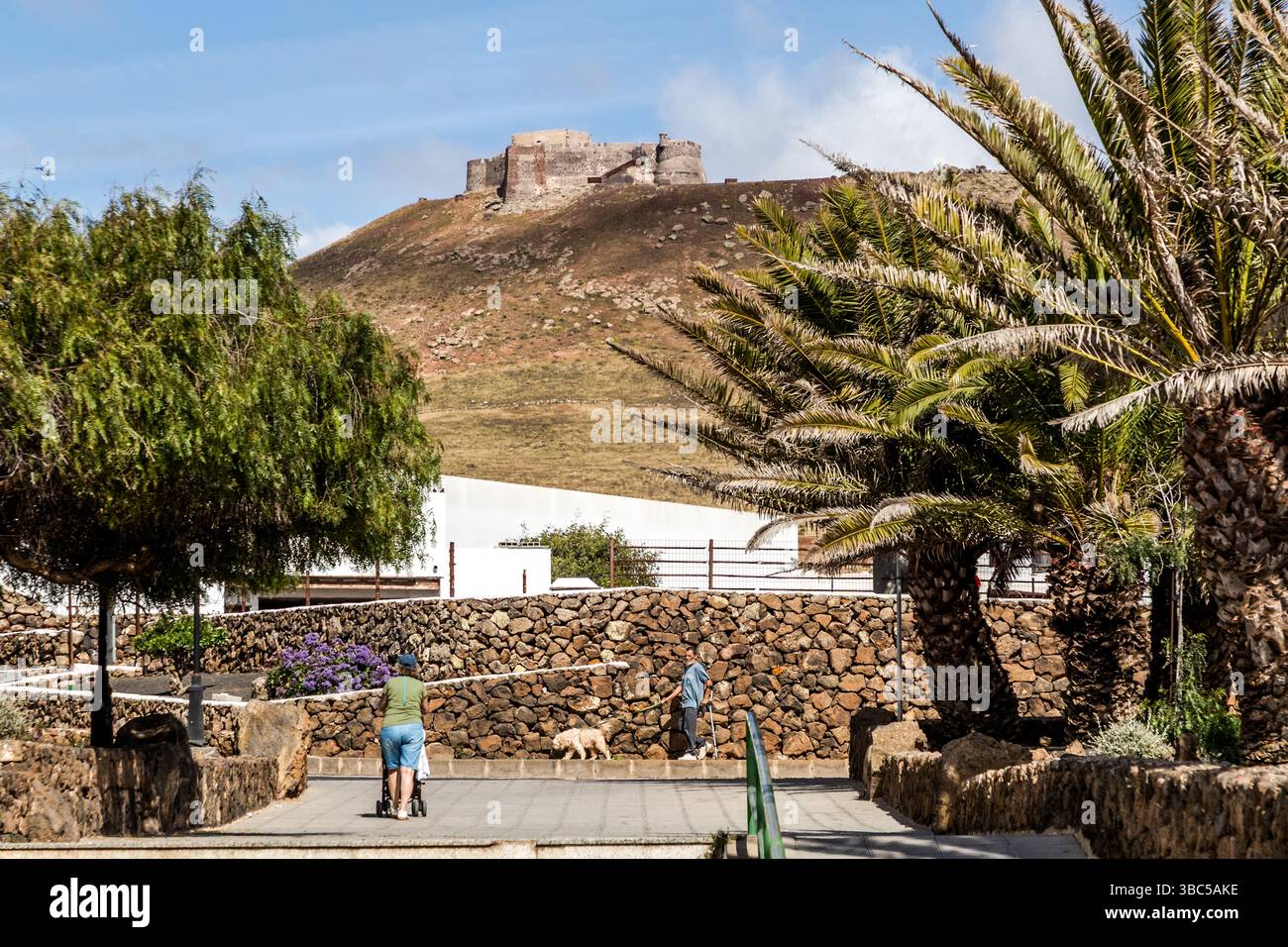 Two people walk through Teguise with the Castillo de Santa Bárbara on ...