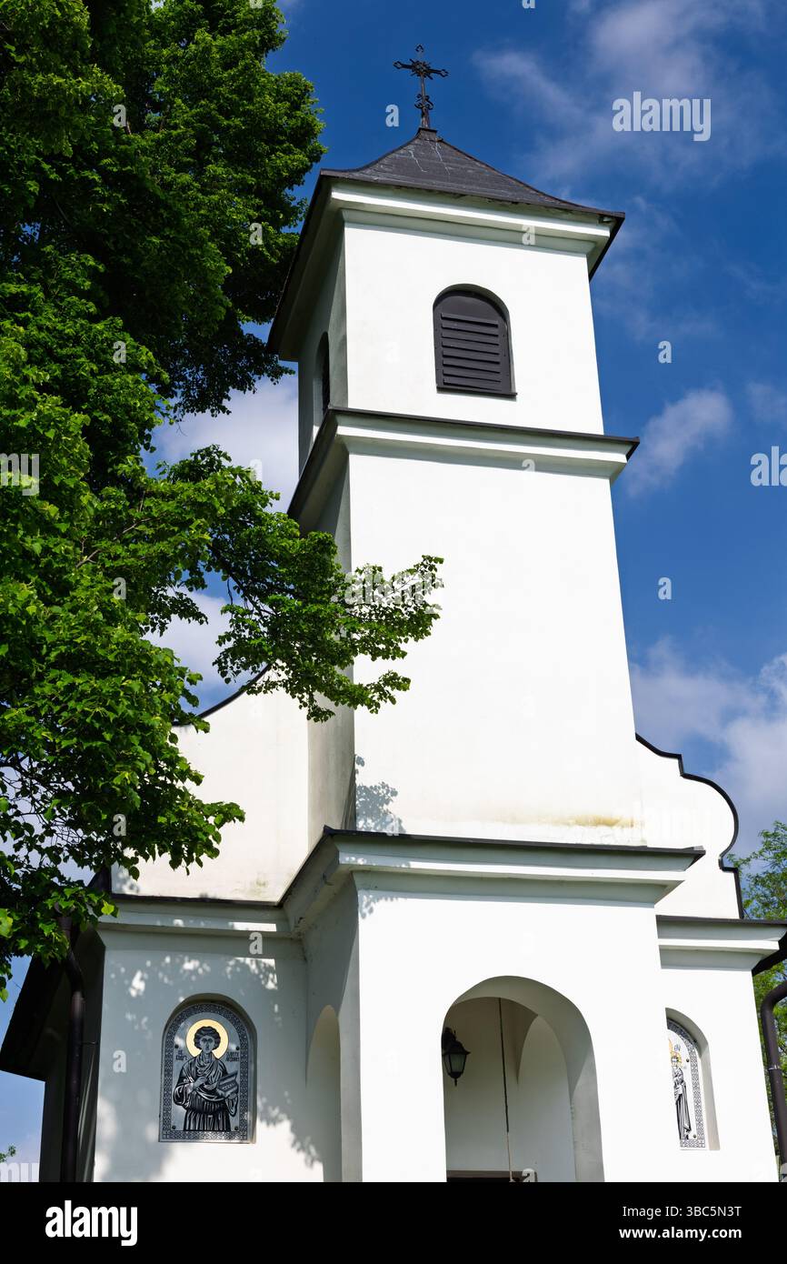 A white church tower with an overhead cross stands surrounded by lush ...
