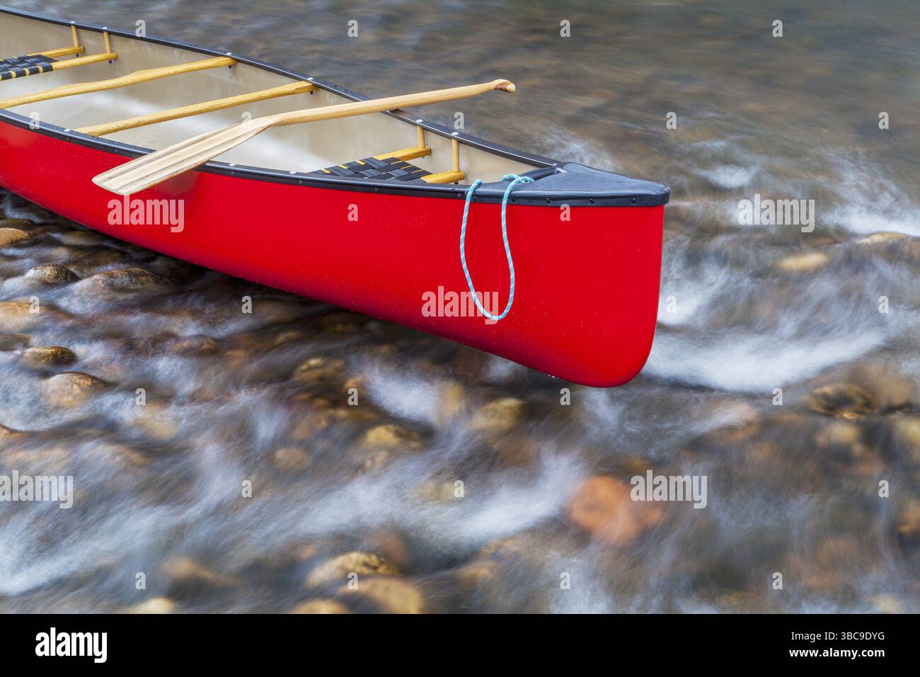 Red canoe bow with a rope and paddle against a shallow river rapid ...